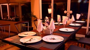 Mesa de jantar posta para quatro, com cadeiras, uma planta e vista pela janela.