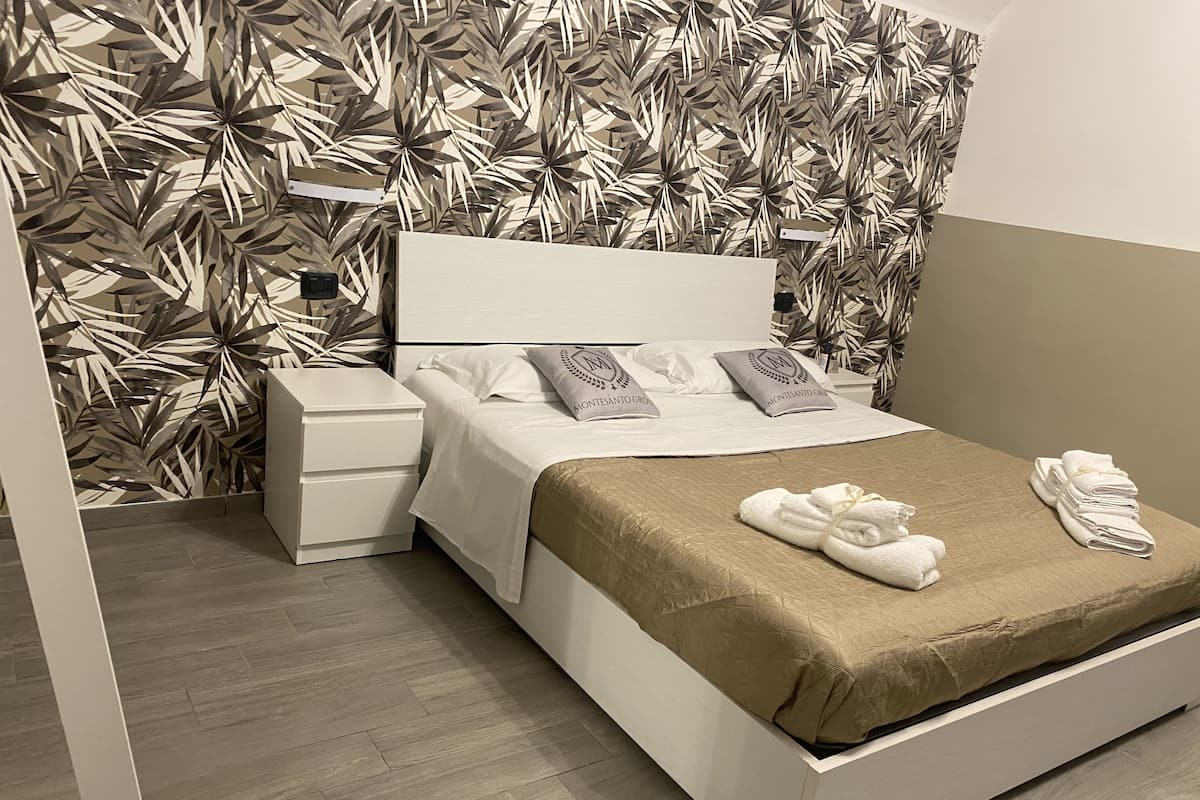 Uma cama bem arrumada com um cobertor marrom e duas almofadas, uma mesa de cabeceira com abajur, e uma parede com padrão de folhas.