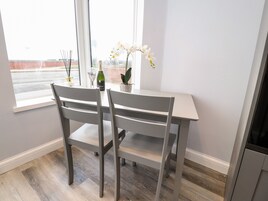 Uma área de refeições com uma mesa, duas cadeiras, uma janela com vista para a rua e um vaso de planta sobre a mesa.