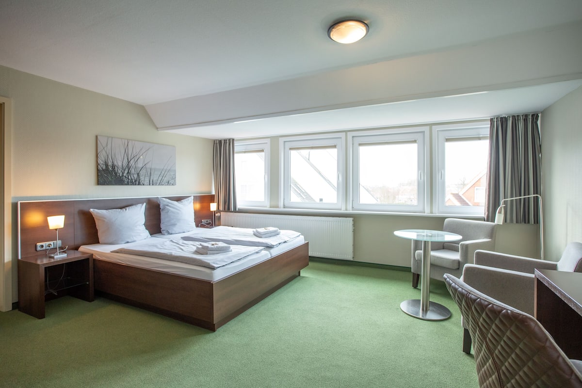 Quarto de hotel com cama, mesas de cabeceira, escrivaninha, cadeira e janela com cortinas.