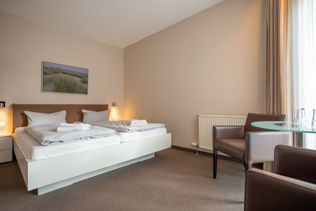 Quarto de hotel com cama, mesas de cabeceira, uma mesinha de vidro e uma cadeira.