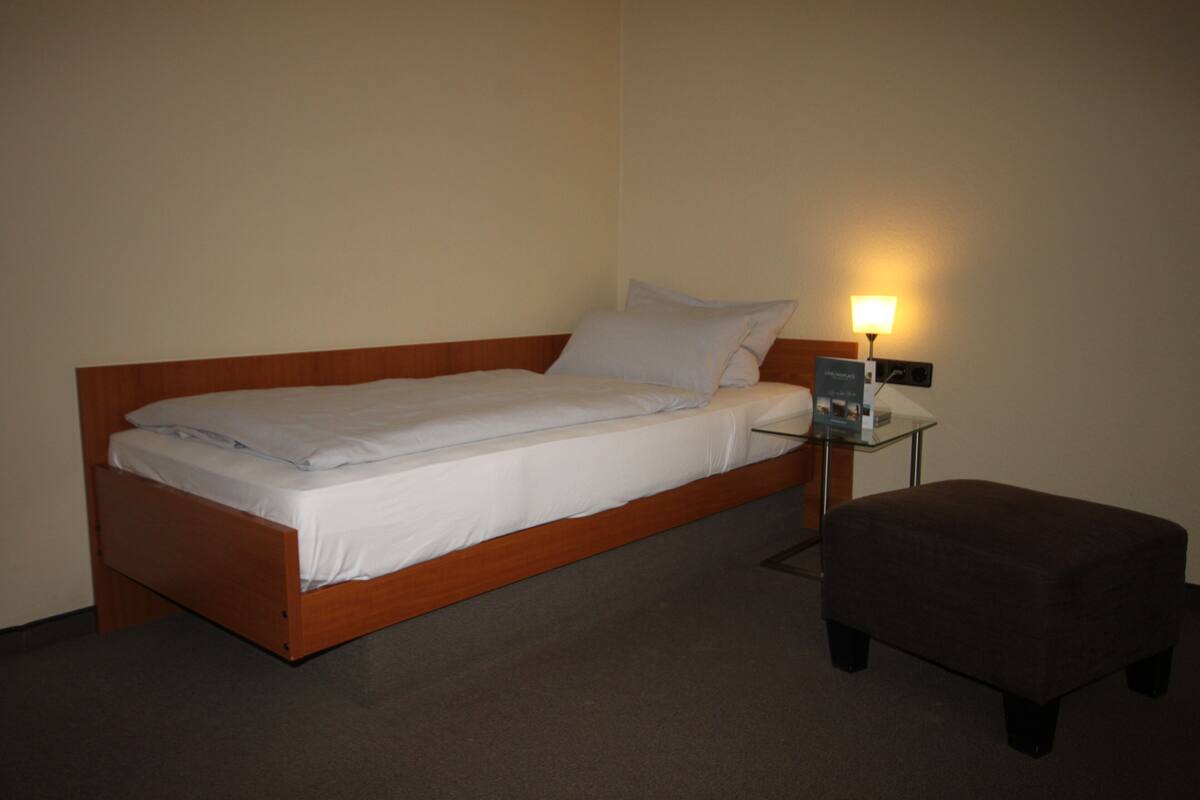 Uma cama de solteiro com cabeceira de madeira, uma mesinha de cabeceira com abajur e um pequeno banquinho.