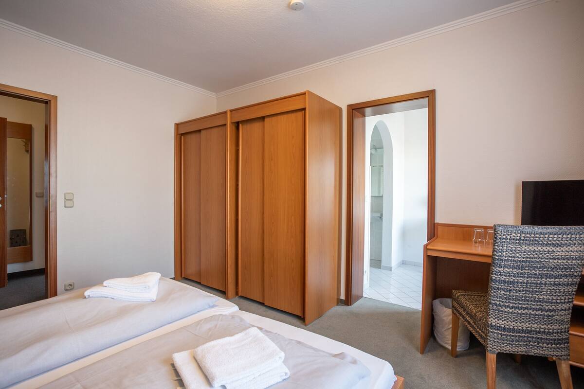 Quarto de hotel com duas camas, um guarda-roupa de madeira, uma escrivaninha com cadeira e uma televisão.
