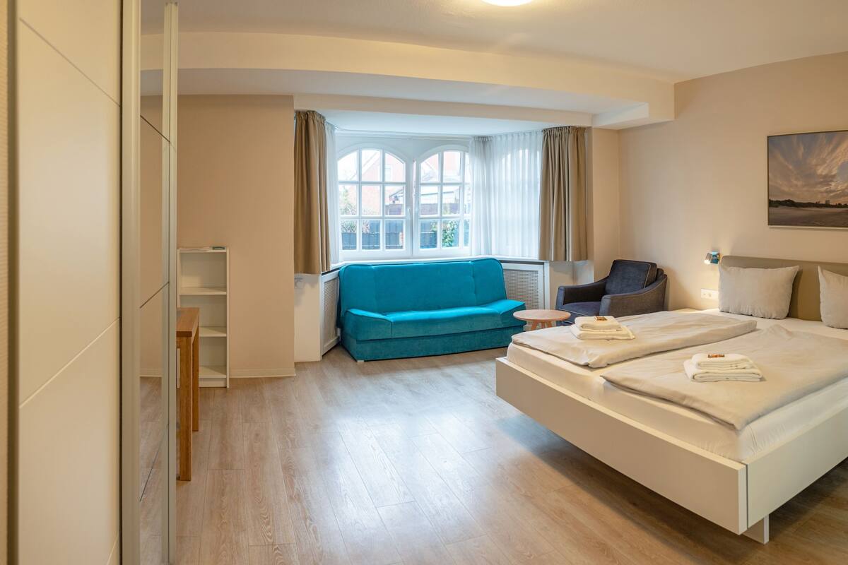 Quarto de hotel moderno com uma cama grande, um sofá azul, uma mesinha e uma janela com cortinas.