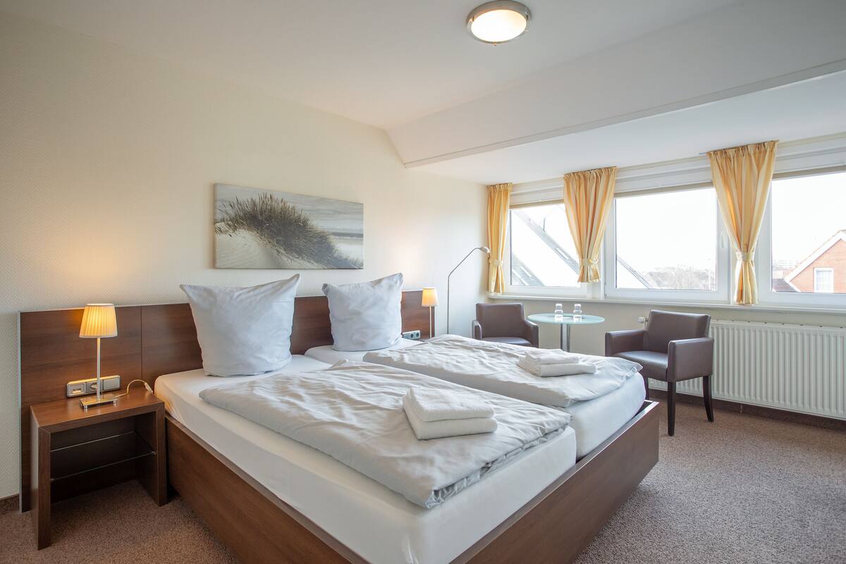 Quarto de hotel com uma cama grande, mesas de cabeceira, luminária, cadeira, mesinha e janela com cortinas.