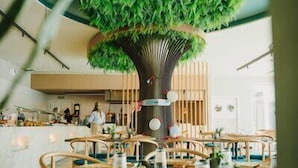Restaurante com mesas redondas, uma planta verde no centro e um design de teto moderno.