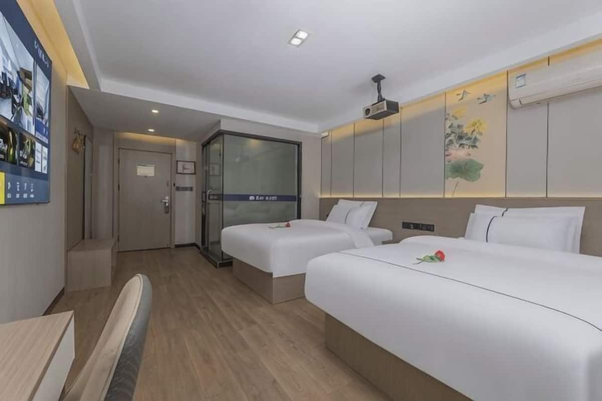 Quarto de hotel com duas camas, uma escrivaninha, divisória de vidro e televisão presa à parede.