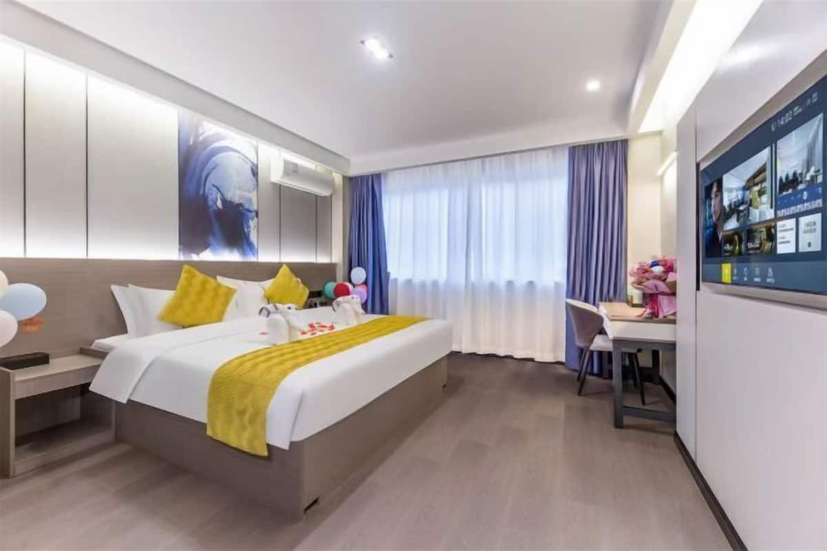 Quarto de hotel com cama, mesa de cabeceira, escrivaninha e uma janela grande com cortinas.
