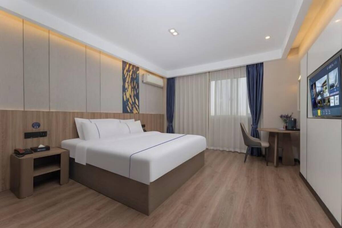 Quarto de hotel com cama, escrivaninha, cadeira, televisão e janela com cortinas.