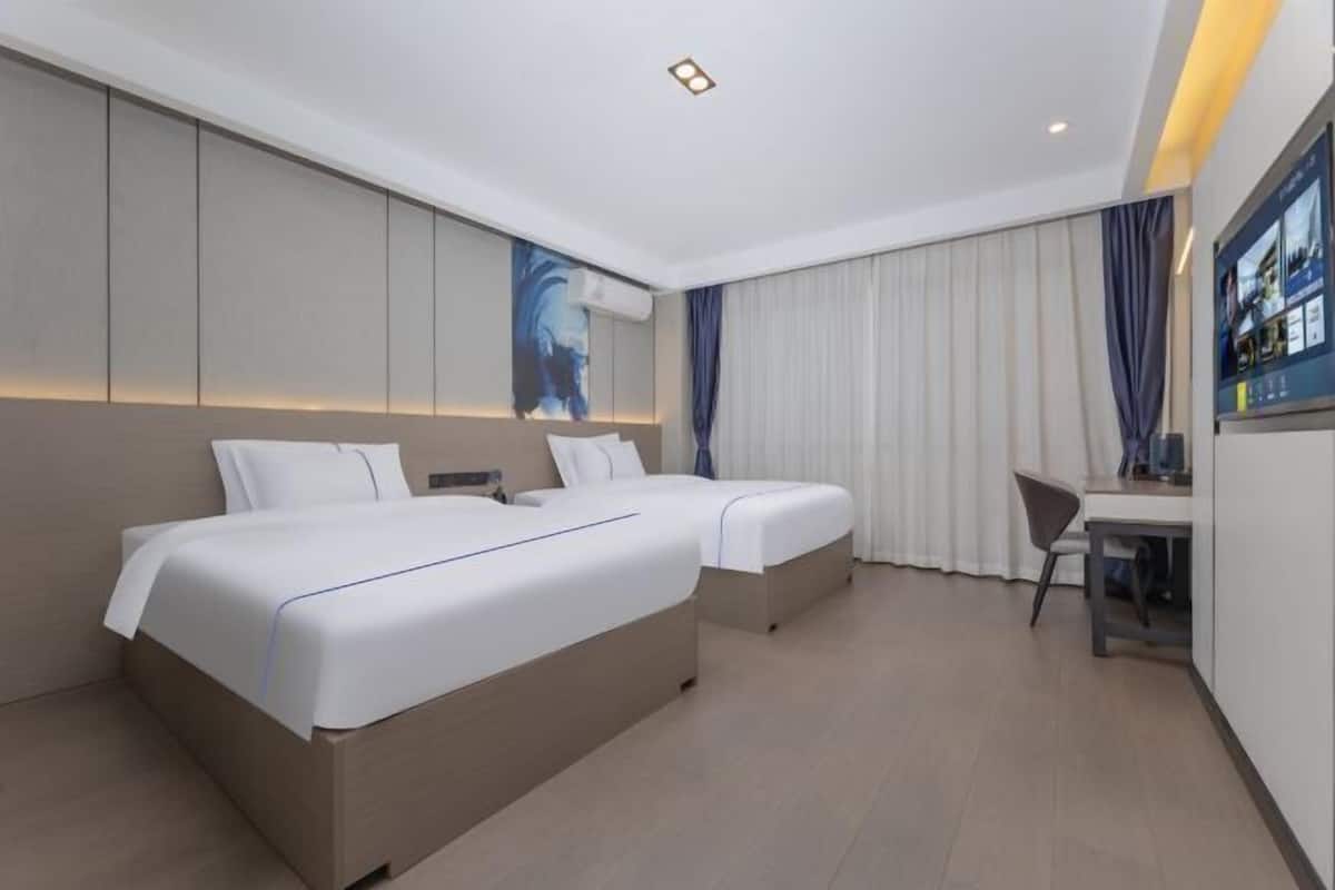 Quarto de hotel moderno com duas camas, uma escrivaninha e uma janela grande com cortinas.