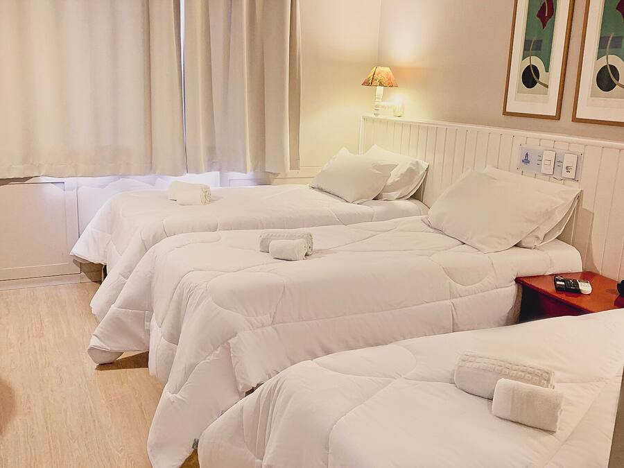 Quarto de hotel com três camas, um radiador, uma luminária e quadros nas paredes.