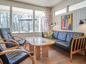 Um quarto aconchegante com piso de madeira, um sofá de couro, duas poltronas de couro, uma pequena mesa de madeira e uma janela com vista para árvores.