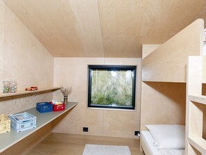 Quarto com cama de solteiro, janela com vista para árvores, prateleira com brinquedos e recipientes, e tapete branco.