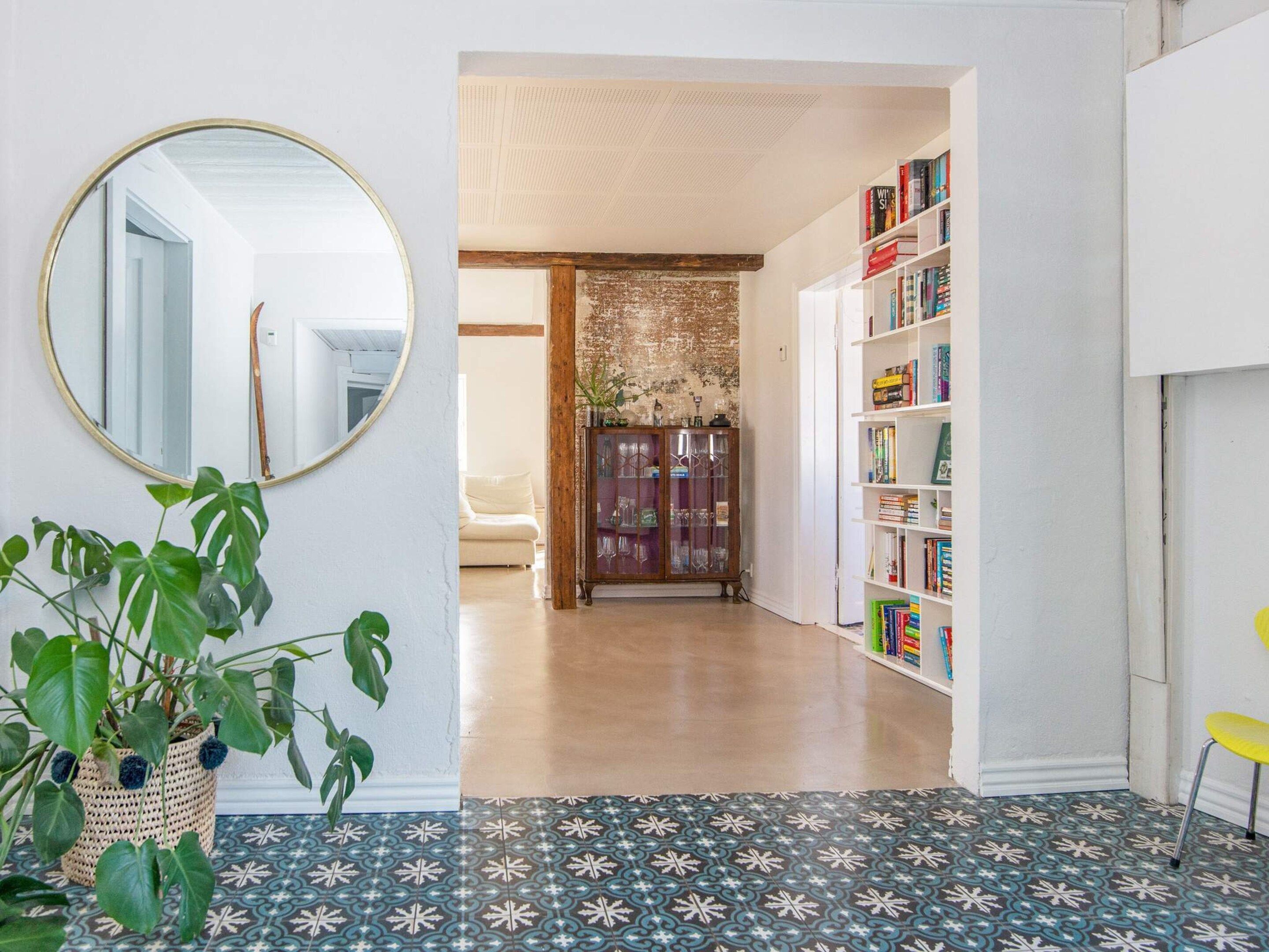 Um corredor com espelho, planta em vaso, estante de livros e piso estampado.