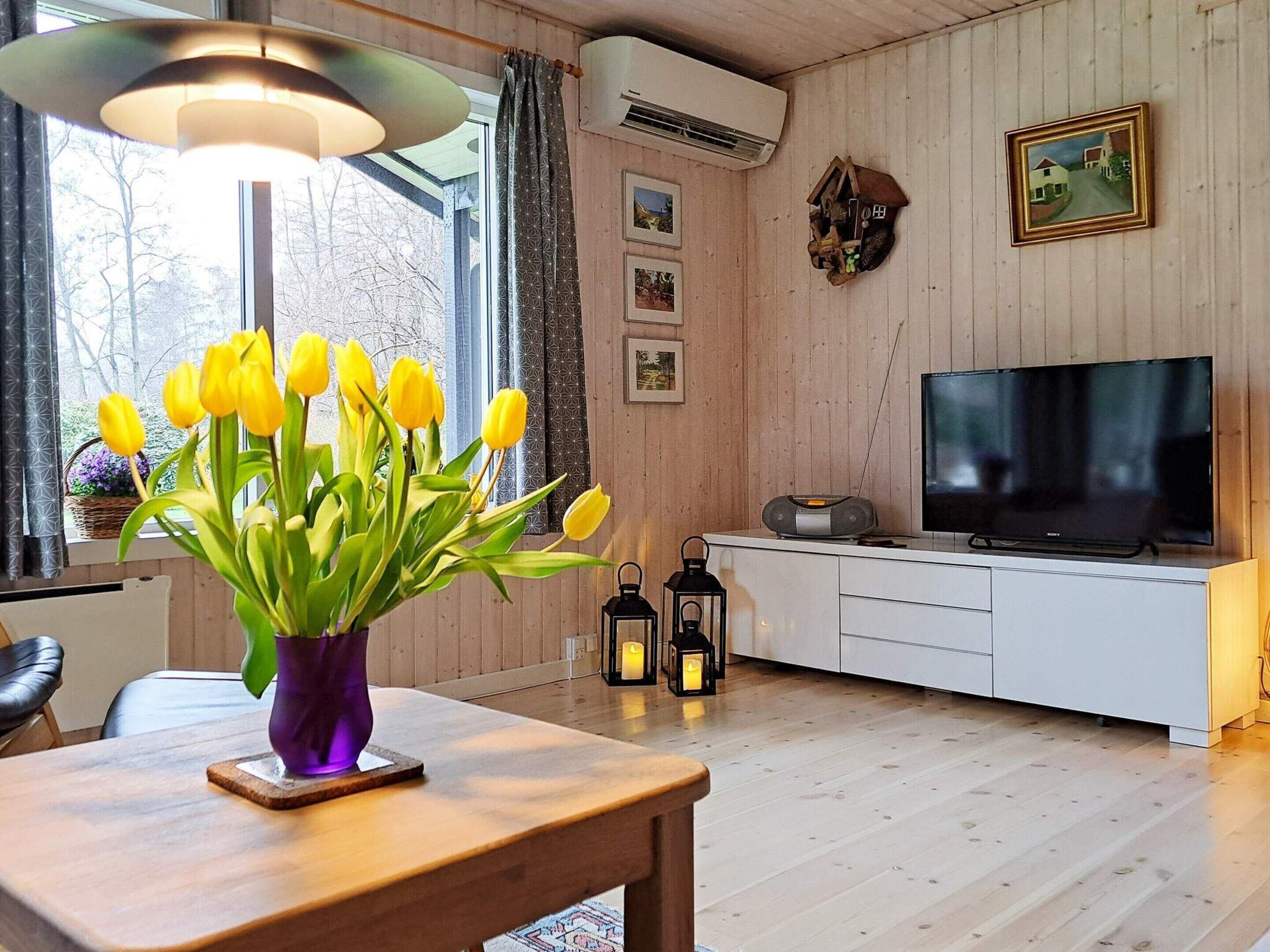 Uma sala de estar com piso de madeira, um suporte branco para TV, uma TV grande, um vaso com tulipas amarelas e um lustre.