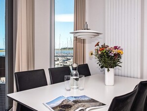 Uma sala de jantar com uma mesa posta para dois, um vaso de flores e vista para um porto através de grandes janelas.