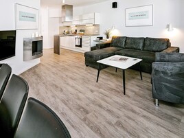 Uma sala de estar moderna com um sofá cinzento, uma mesa de centro e uma televisão. Uma cozinha visível ao fundo possui armários brancos e um fogão.