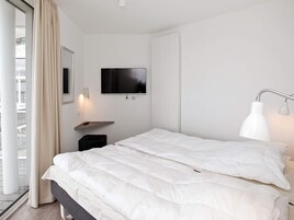 Quarto de hotel moderno com uma cama grande, televisão fixada na parede, mesa de cabeceira e janela com cortinas.