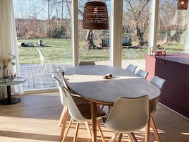 Uma área de jantar moderna com uma grande mesa redonda, cadeiras brancas e vista para um jardim e árvores através de amplas janelas.