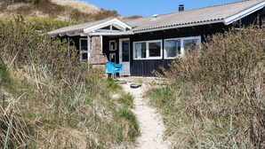 Uma casa costeira com um caminho de areia que leva à entrada, rodeada por vegetação densa.