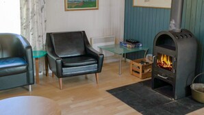 Uma sala de estar com uma poltrona de couro preto, uma mesa de vidro, um caixote de madeira e uma lareira a lenha.
