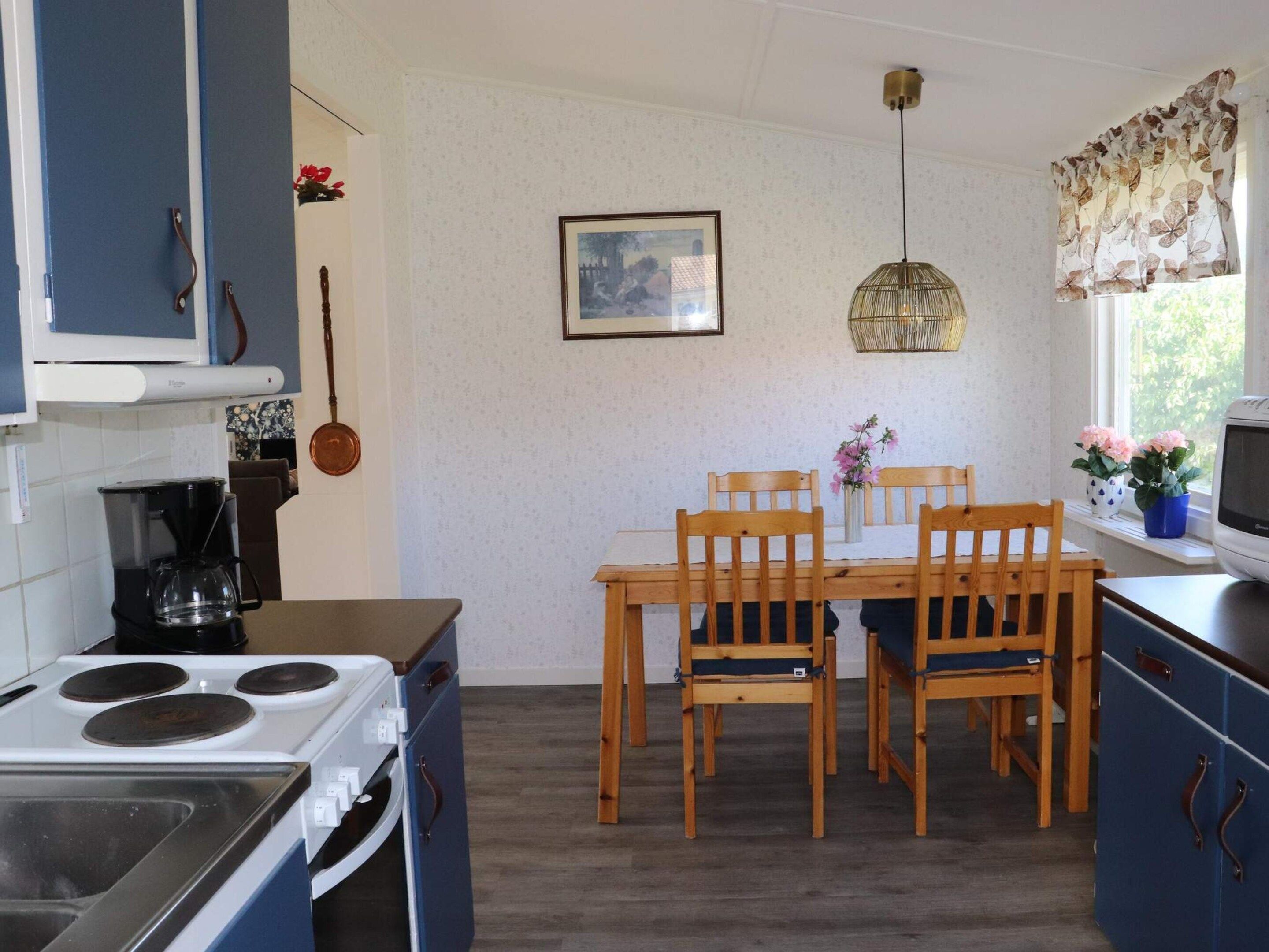 Cozinha compacta com fogão, micro-ondas e cafeteira. Há uma mesa de jantar com cadeiras e uma janela com cortinas.