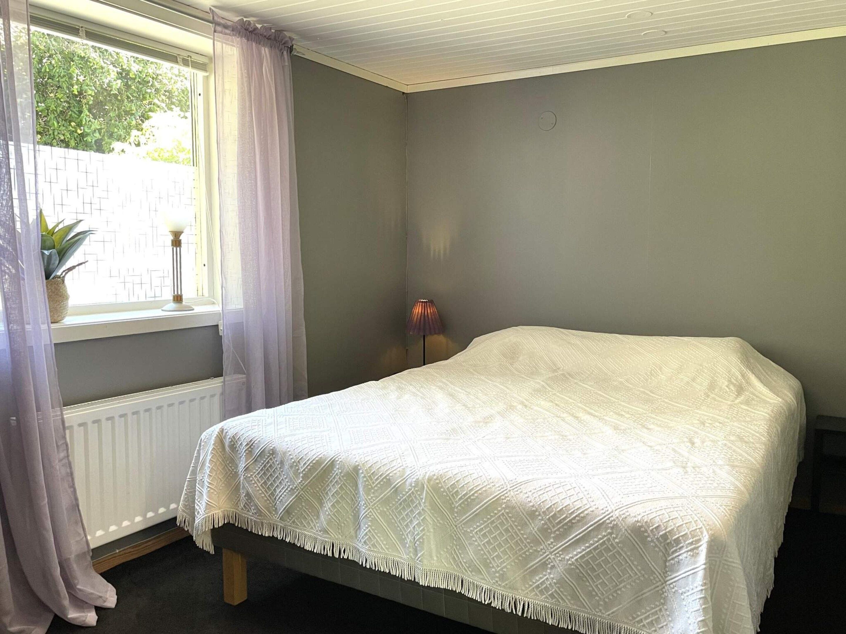 Quarto com uma cama de solteiro, janela com cortinas, radiador e uma pequena planta no parapeito.