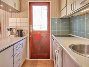 Uma cozinha compacta com armários de madeira, cafeteira, suporte de panela vermelho, máquina de lavar e painel de azulejos azul.