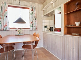 Uma área de refeições com mesa de madeira, cadeiras, luminária pendente, janela com cortinas e uma cozinha visível por uma porta aberta.