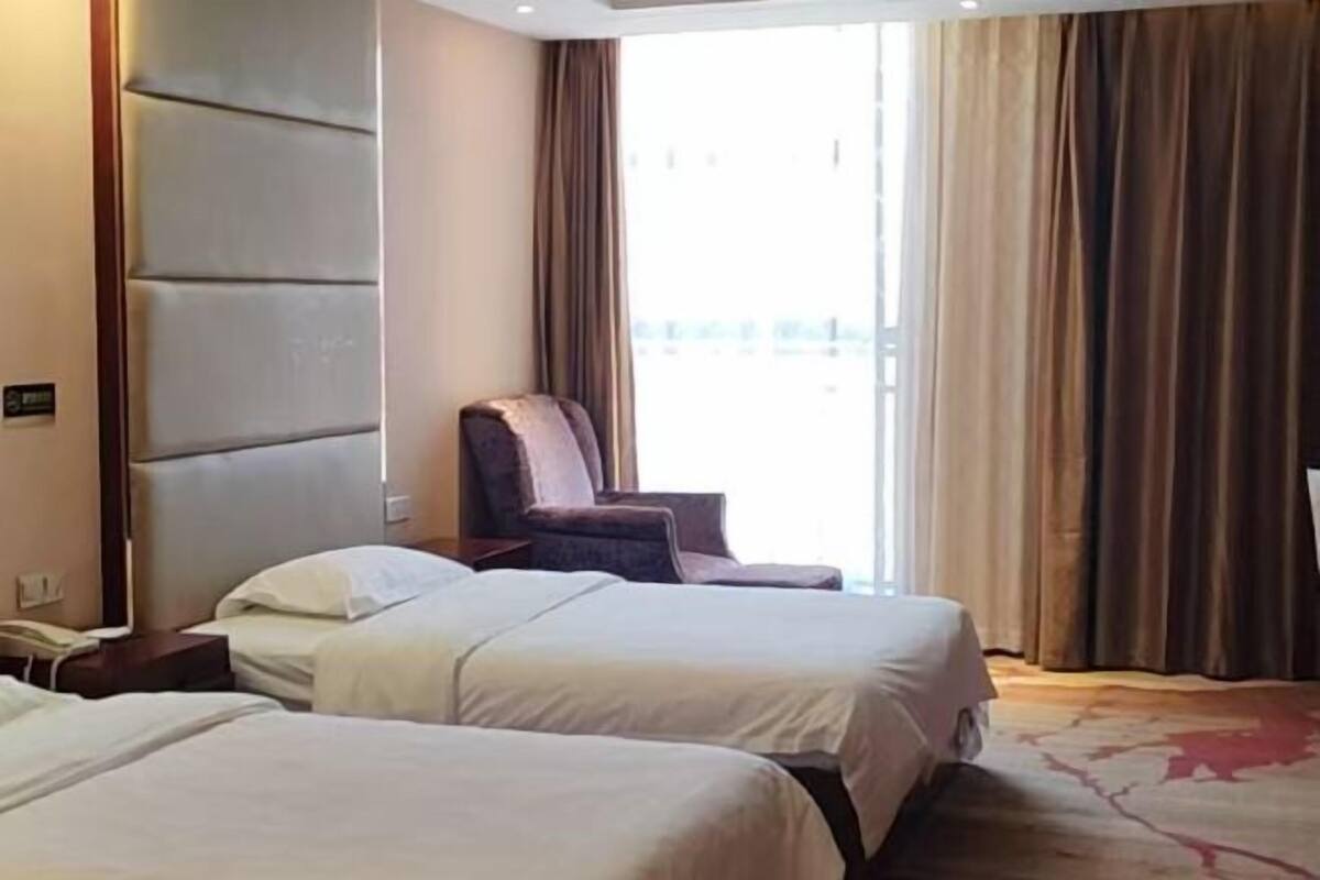 Quarto de hotel com duas camas, uma cadeira, uma escrivaninha com telefone e uma janela grande com cortinas.