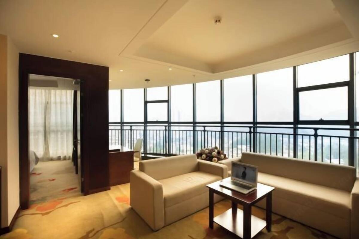 Quarto espaçoso com uma grande janela que oferece vista para a cidade, um sofá, uma mesinha com um laptop e uma planta decorativa.