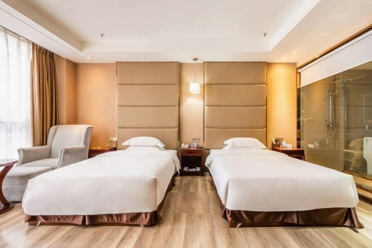 Quarto de hotel com duas camas, área de estar com uma poltrona, uma janela grande e chuveiro de vidro.