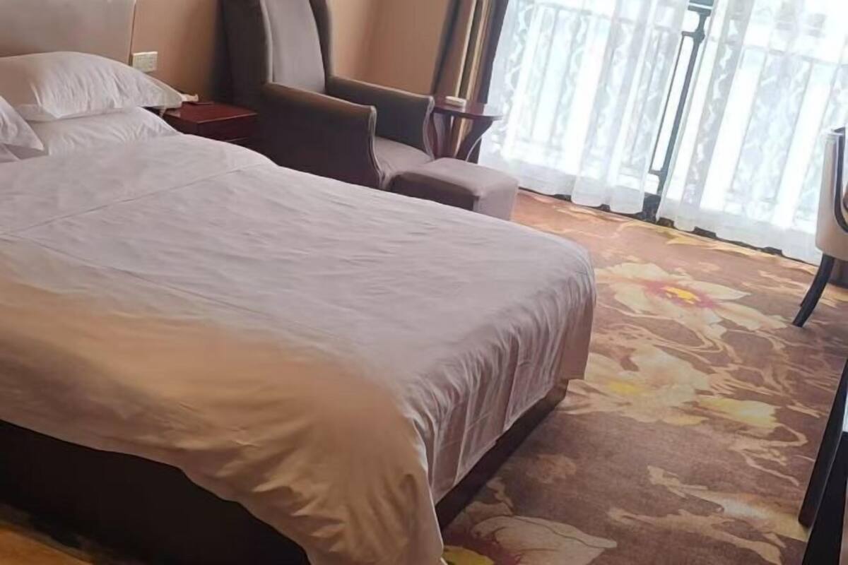Quarto de hotel com uma cama grande, uma cadeira, uma mesa de cabeceira e uma janela com cortinas.