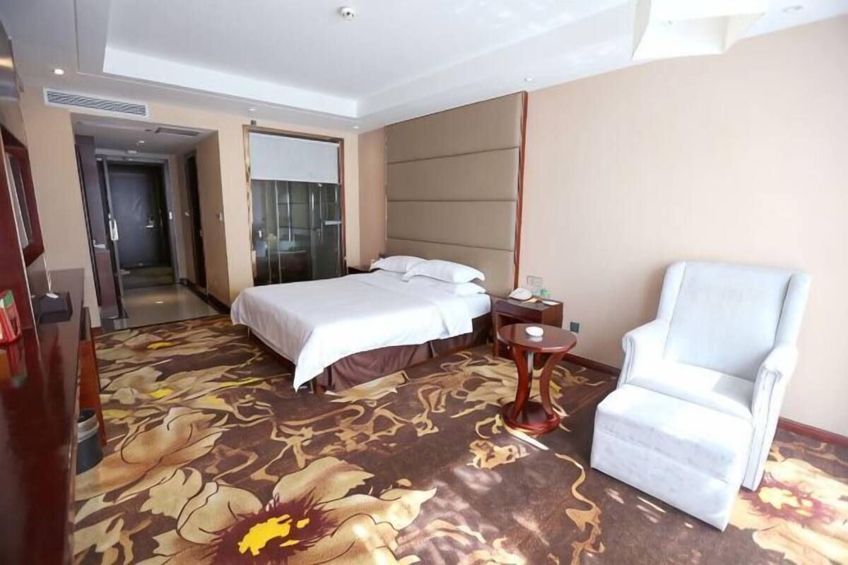 Quarto de hotel com uma cama, uma cadeira, uma mesinha e uma porta que dá para outro cômodo.