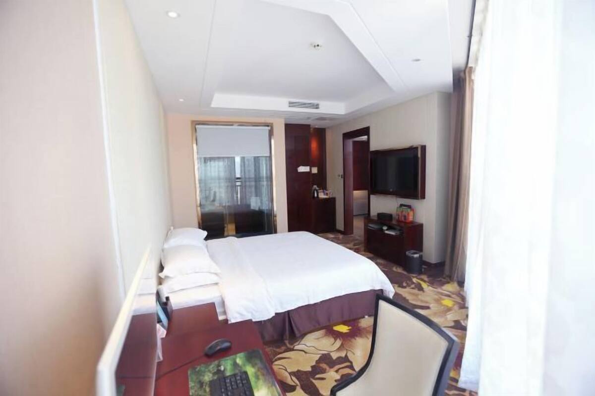 Quarto de hotel com uma cama, uma escrivaninha com computador, uma cadeira, uma televisão e uma janela com cortinas.