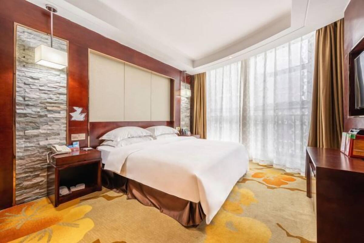 Quarto de hotel com cama, mesa de cabeceira, abajur, janela grande com cortinas e parede de pedra decorativa.