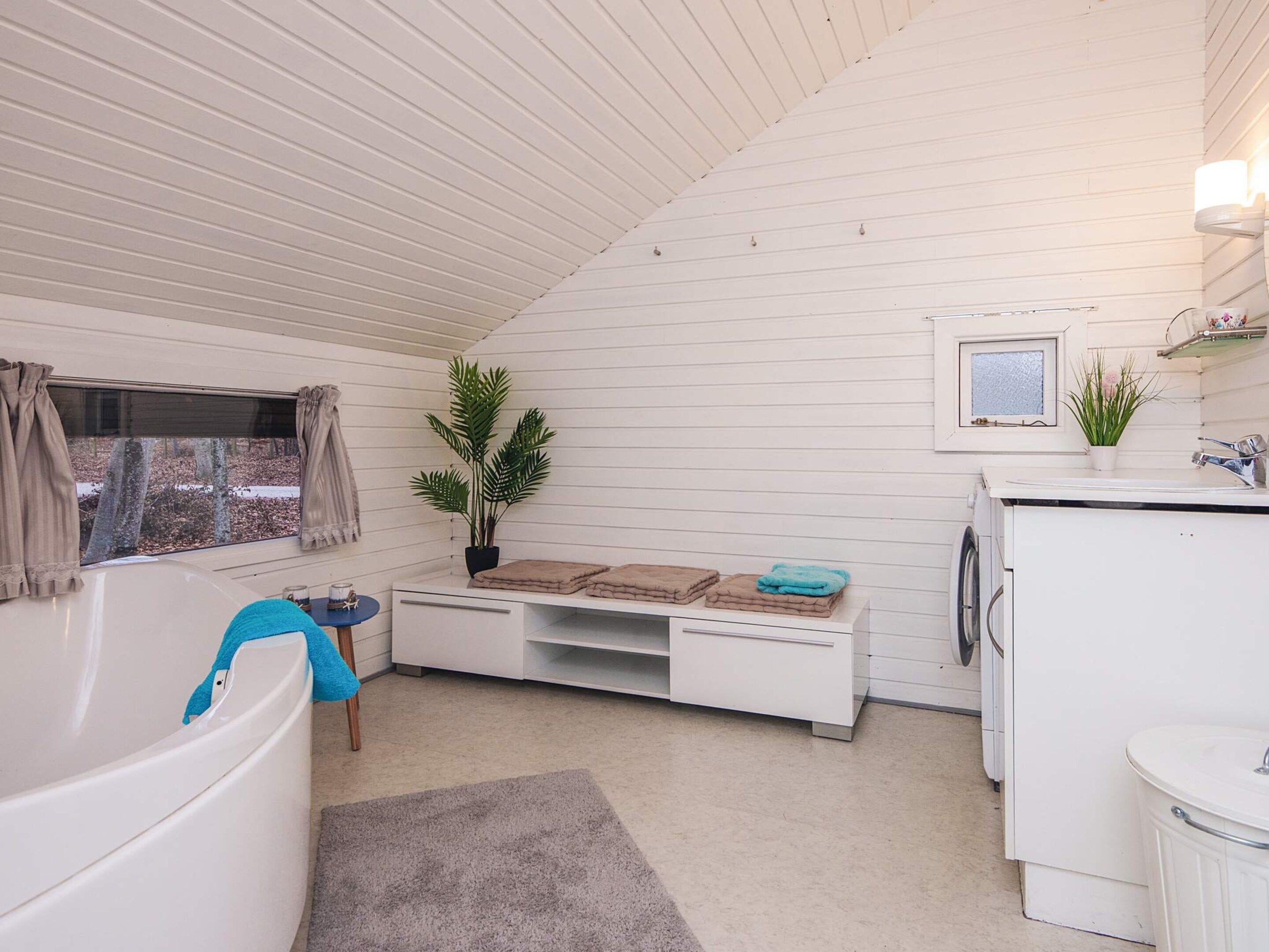 Casa de banho compacta com decoração em branco, banheira, pia, pequena janela e um vaso de planta.