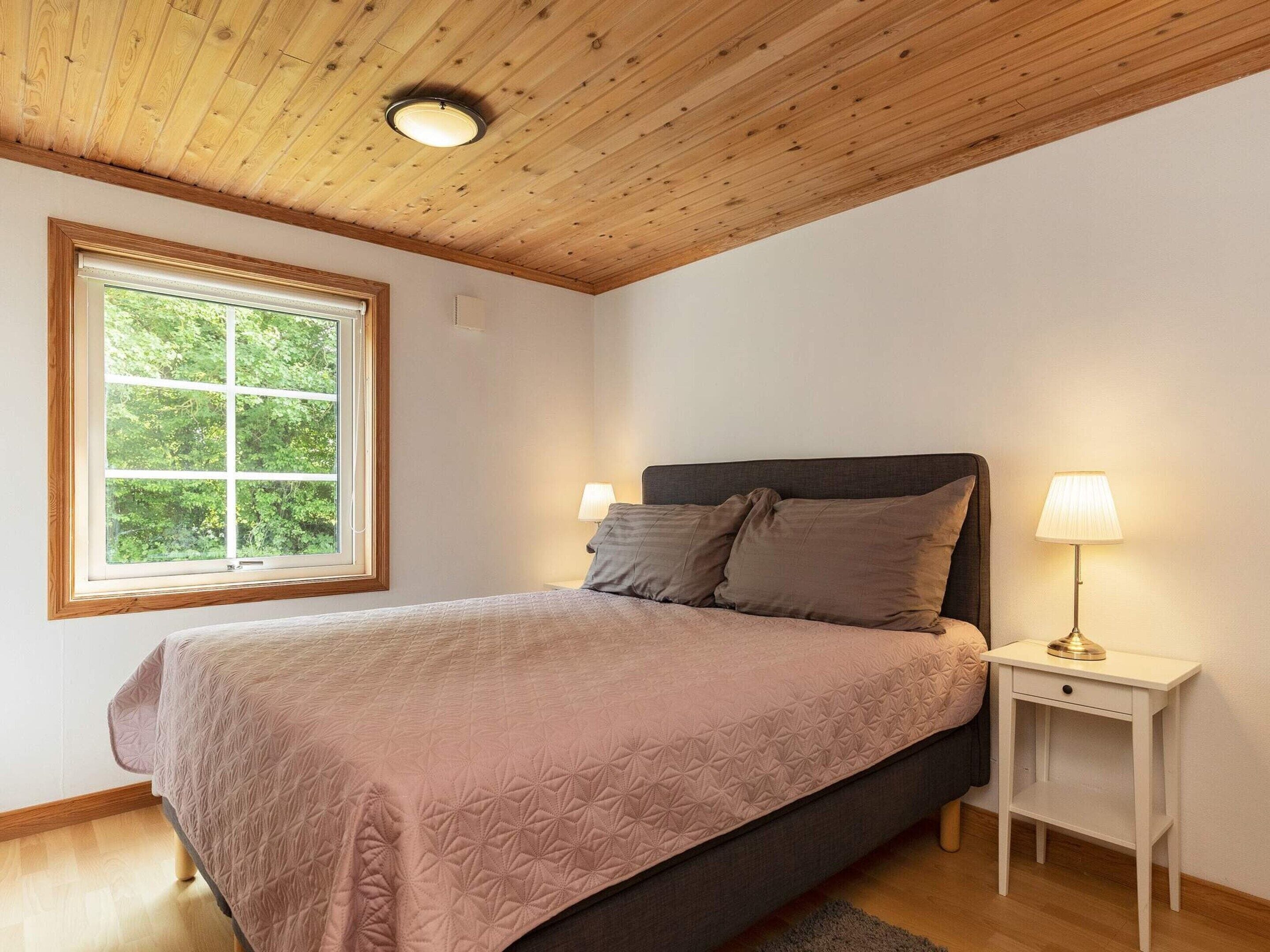 Quarto com cama, mesa de cabeceira com abajur, janela com vista para árvores e teto de madeira.