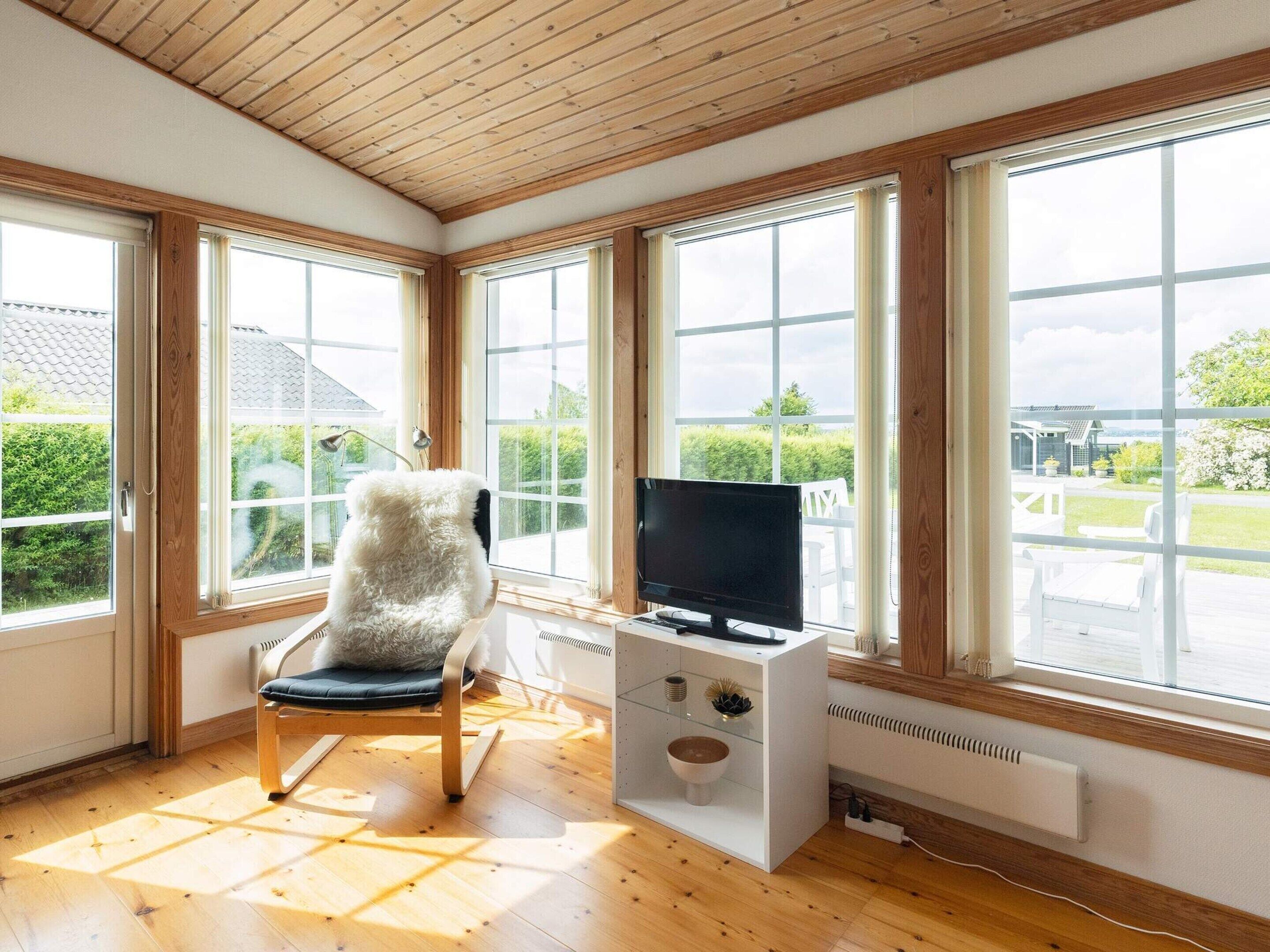 Quarto com amplas janelas, televisão sobre um suporte, uma cadeira e um radiador.