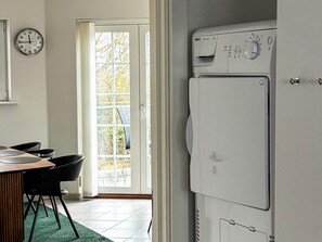 Quarto com uma janela grande, relógio na parede, máquina de lavar e área de jantar com cadeiras e mesa.
