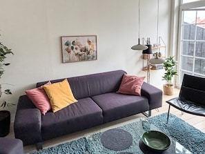 Uma sala de estar moderna com um sofá cinza escuro, duas almofadas, uma obra de arte floral emoldurada, um abajur, uma mesa lateral com livros, uma planta em vaso e uma janela com vista.