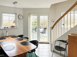 Uma área de refeições com mesa de madeira, cadeiras pretas, um relógio e vista para um pátio externo através de uma porta de vidro.