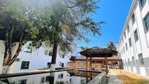 Um pátio com um pavilhão tradicional, um espelho d'água e edifícios modernos ao fundo.