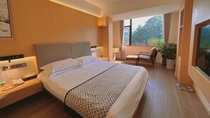 Quarto de hotel com cama, mesa de cabeceira, cadeira, planta em vaso e janela com vista para vegetação.