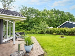 Um deck de madeira com móveis de exterior, uma planta em vaso e uma pequena área de jardim com relvado e árvores.