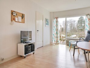 Quarto com televisão sobre carrinho, mesa de jantar com cadeiras, porta e vista para o exterior.