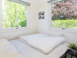 Quarto com cama, duas janelas, uma pequena mesa com planta e tomada na parede.