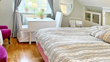 Quarto com cama grande, escrivaninha com cadeira, mesinha com plantas e janela com cortinas.