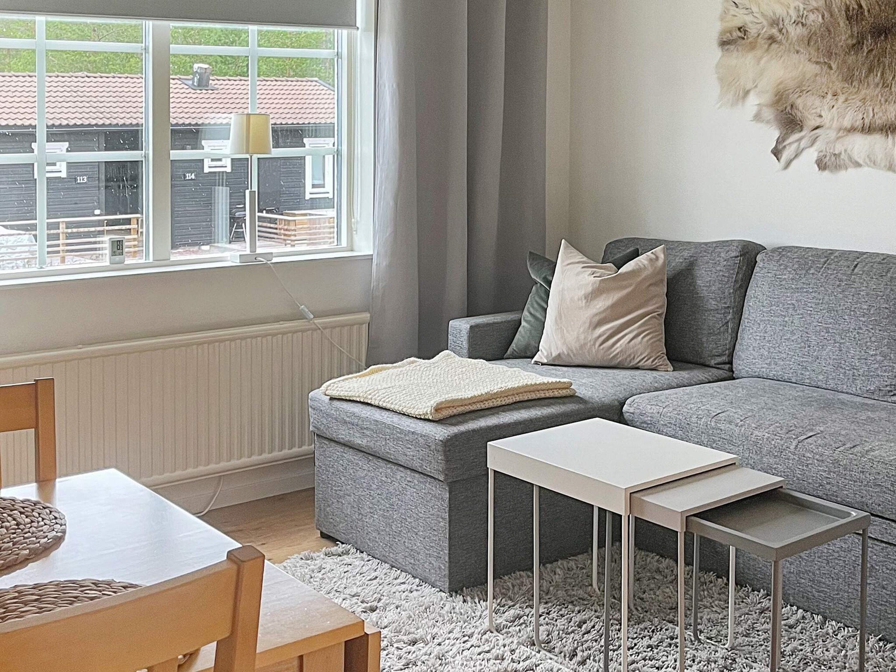 Uma sala de estar moderna com um sofá cinzento, uma mesa pequena branca e uma janela com cortinas.