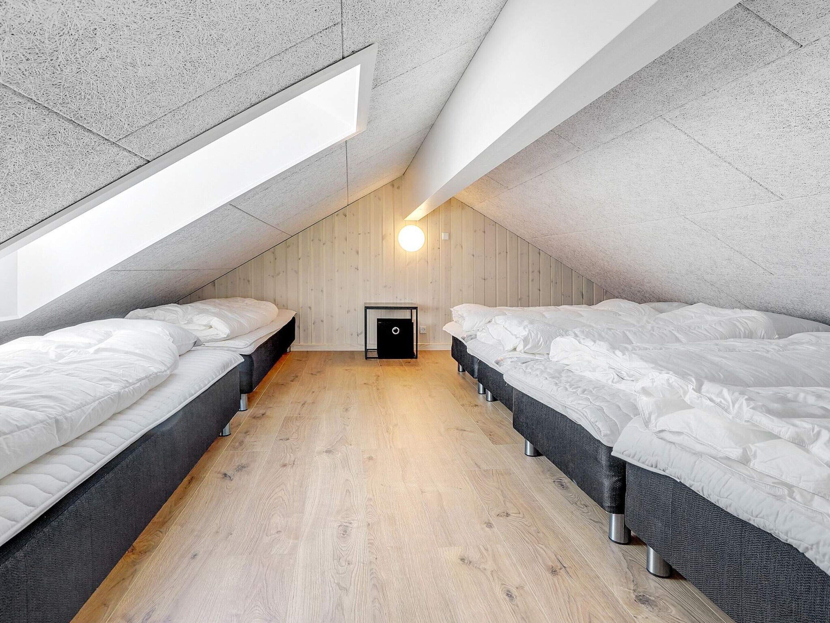 Quarto compacto com teto inclinado, duas camas, uma pequena mesa e luminária.